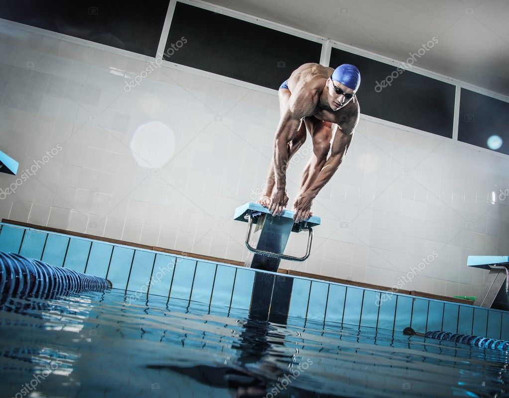 Young muscular swimmer in low position on starting block in a swimming ...