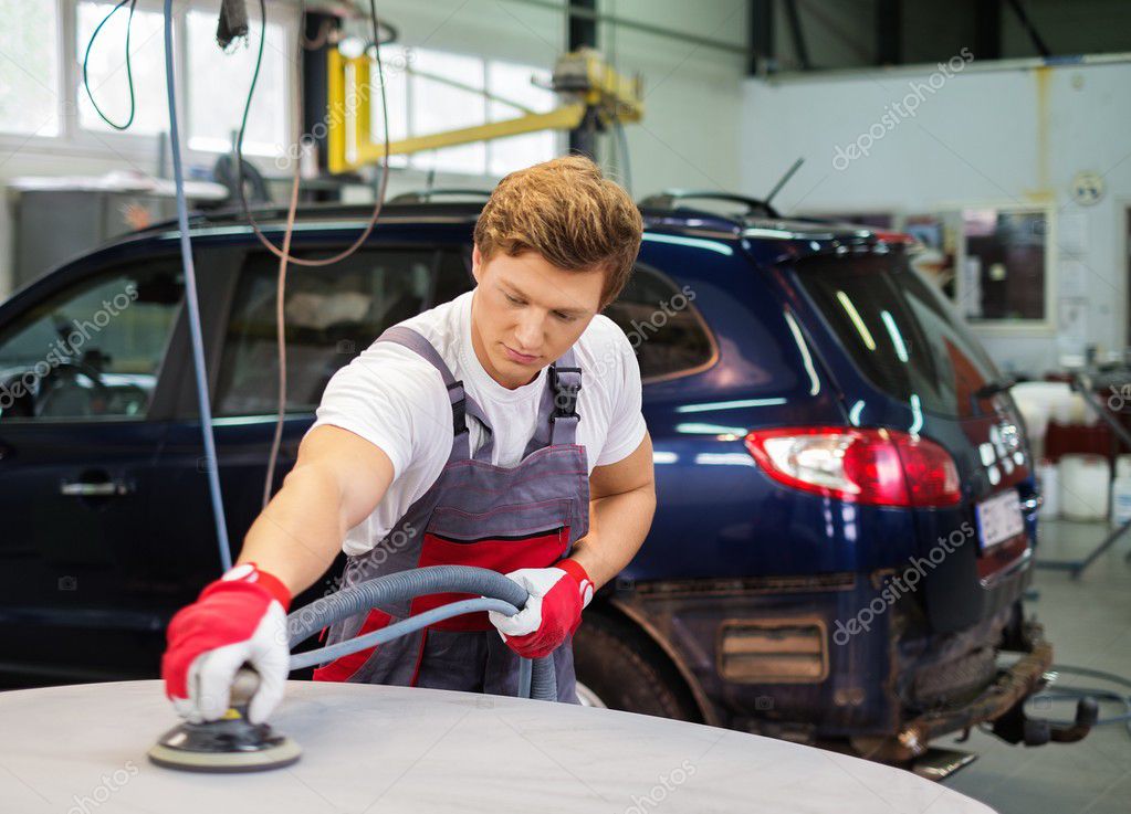 Young serviceman performing grinding with machine on a car bonnet in a ...