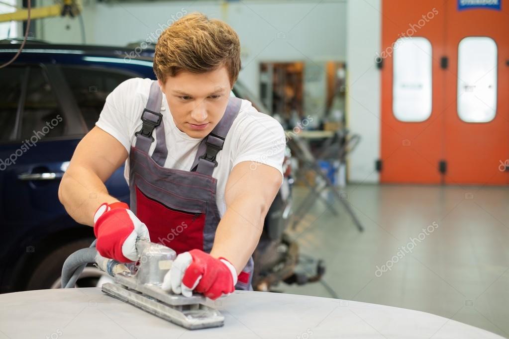 Young serviceman performing grinding with machine on a car bonnet in a ...