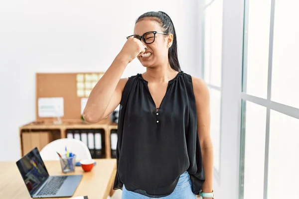 Young hispanic business woman working at the office smelling something ...