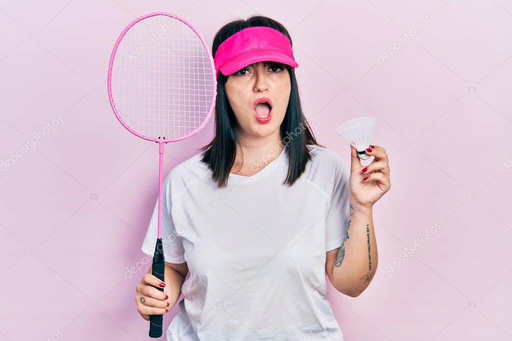 Joven mujer hispana sosteniendo raqueta de bádminton y lanzadera con cara de shock, escéptica y ...
