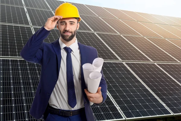 Young male engineer with hardhat and rolls near modern solar power ...
