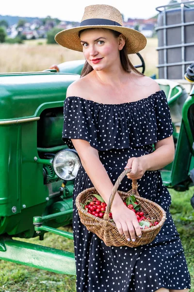 Young woman with a basket of cherries stands near a tractor in the ...