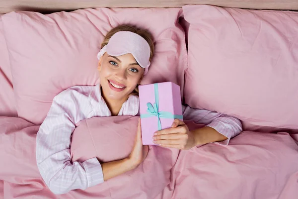 Top view of smiling woman in sleep mask holding present on pink bedding ...