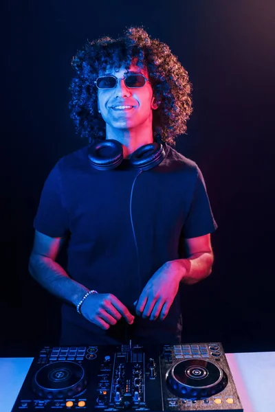 Man with curly hair using DJ equipment and standing in the dark neon ...