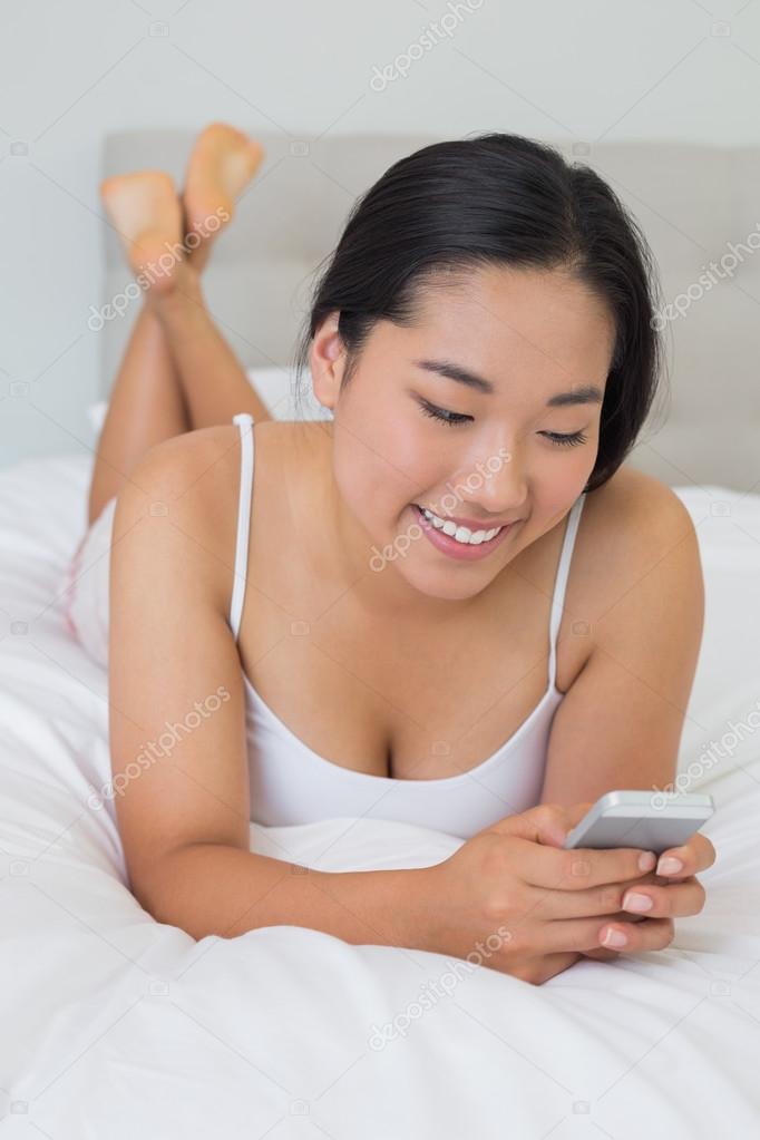 Sonriendo mujer asiática acostada en la cama mensajes de texto en el teléfono: fotografía de ...