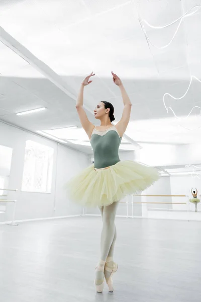 young ballerina in tutu dancing with raised hands during rehearsal in ...