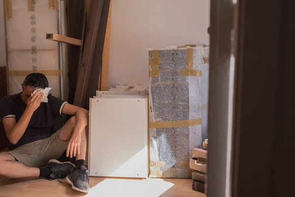 Moving to new home. Tired man sitting on the floor wipes his sweat with ...