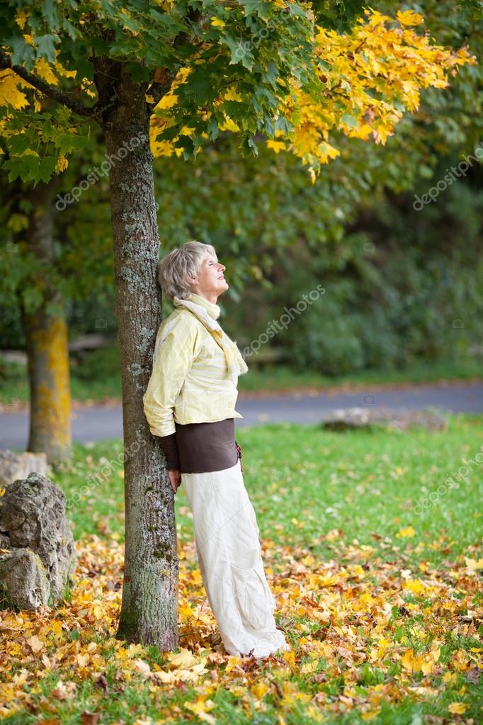 Senior Woman Leaning On Tree Trunk In Autumn At Park Stock Photo by ...
