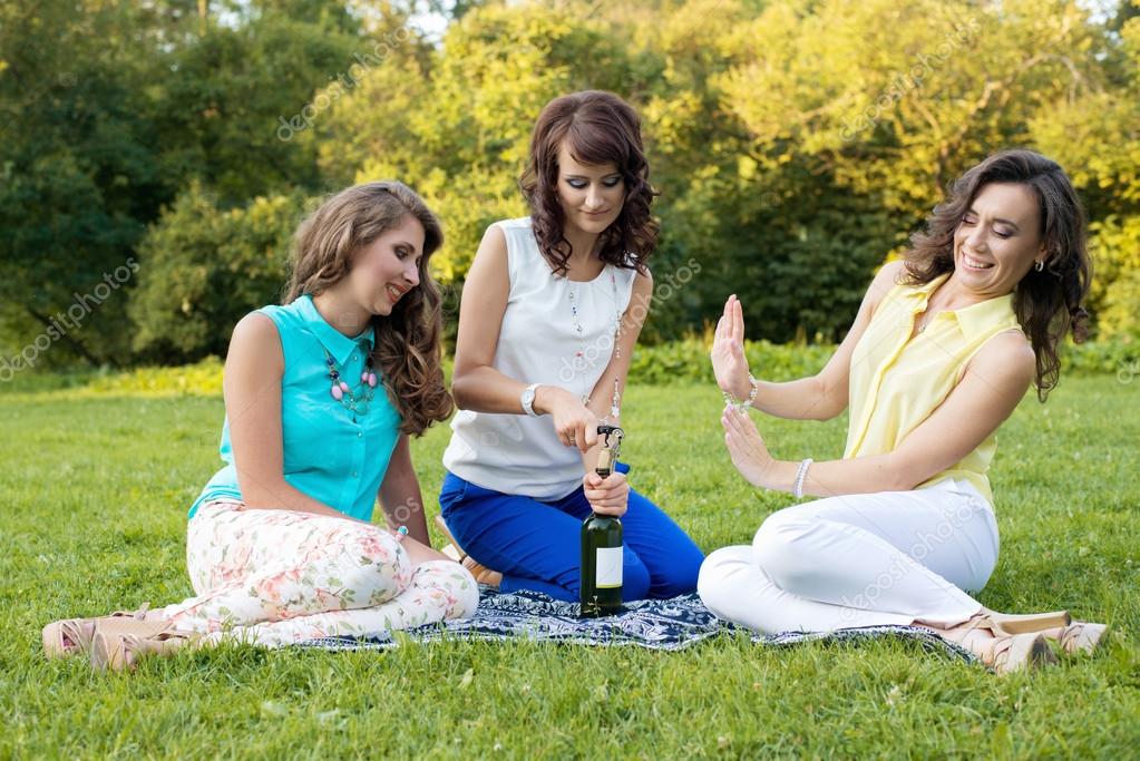 Tres novias guapas jóvenes abriendo una botella de vino en el prado durante el picnic, riendo y ...