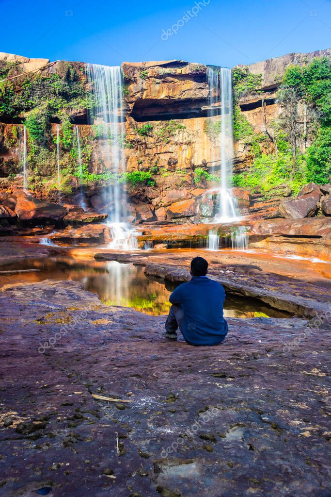 joven aislado sentado cerca de la cascada que cae de la cima de la ...
