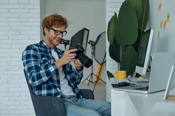 Cheerful young man examining camera while sitting at his working place ...
