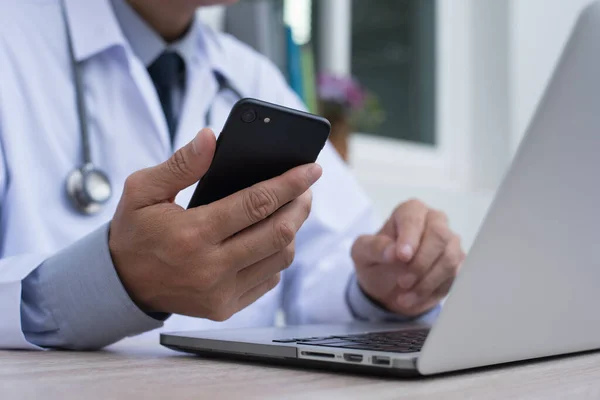 Smiling 30-40s male doctor using smartphone working on laptop computer ...