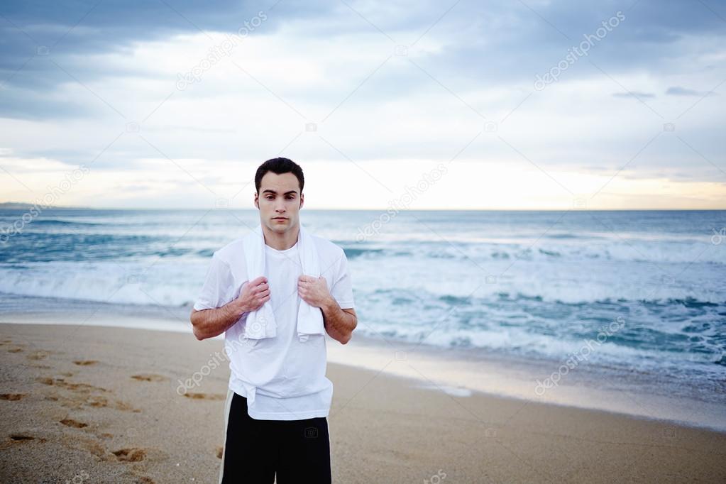 Fit man resting after run while standing on the beach with seas on ...