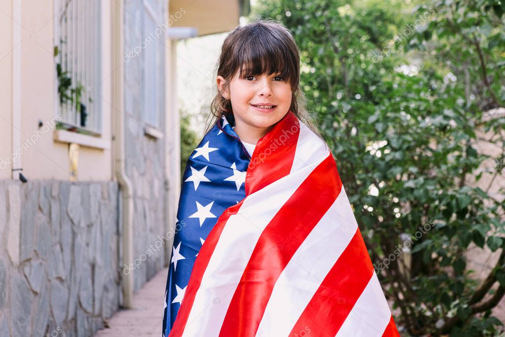Chica de pelo negro cubierta con una gran bandera americana, en el jardín de su casa. Concepto ...
