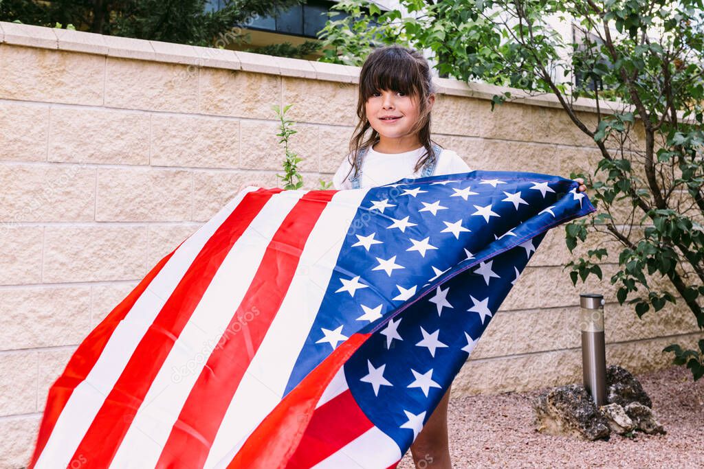 Chica de pelo negro sosteniendo y ondeando una bandera de los Estados Unidos, en el jardín de su ...