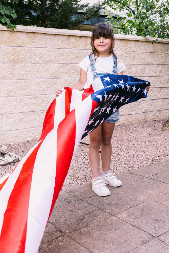 Chica de pelo negro sosteniendo y ondeando una bandera de los Estados Unidos, en el jardín de su ...
