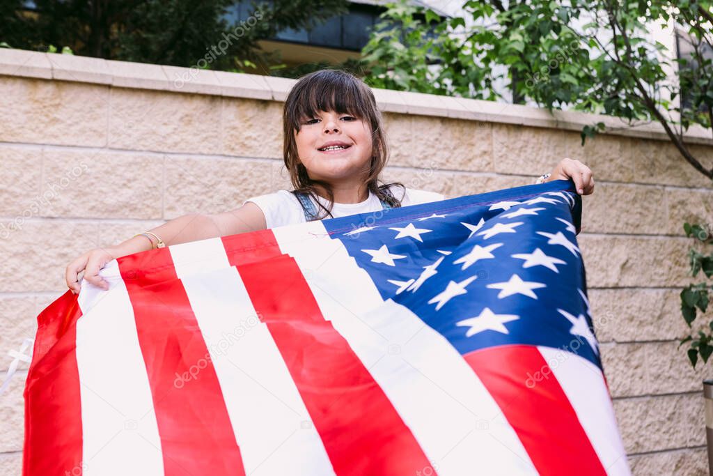 Chica de pelo negro sosteniendo y ondeando una bandera de los Estados Unidos, en el jardín de su ...