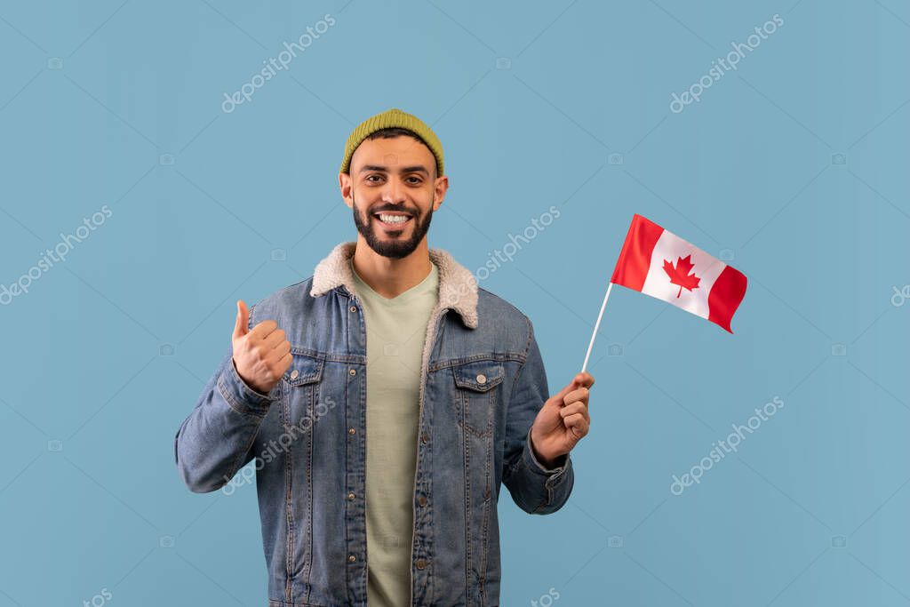 Emocionado hombre de Oriente Medio mostrando el pulgar hacia arriba y la bandera de Canadá, de ...