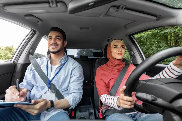 Smiling millennial middle eastern female in hijab at steering wheel driving car taking driving ...