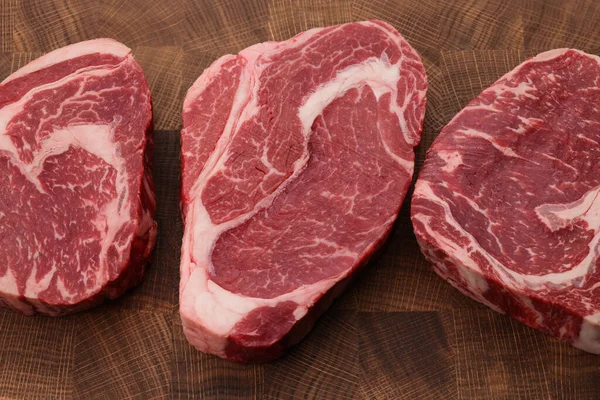 Close up three raw marbled ribeye beef steaks over end grain cutting ...