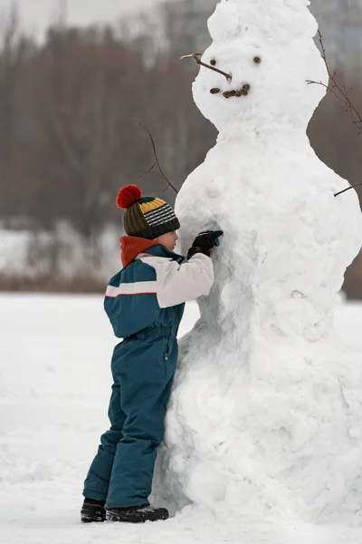 Portrait of little boy and huge snowman. Child sculpts snowman on snowy ...