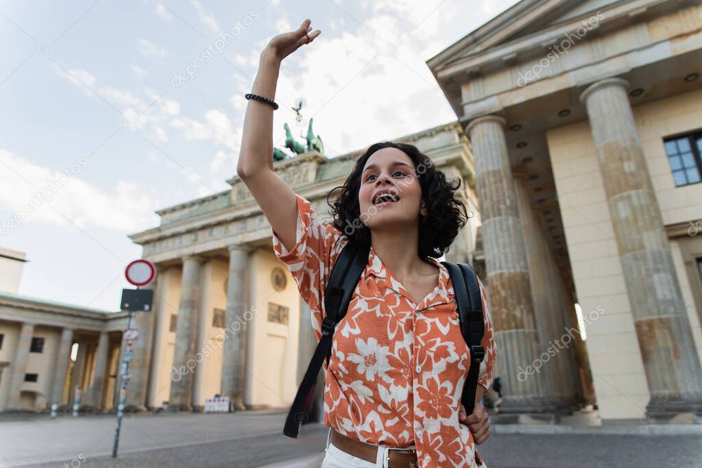 curly tourist with backpack waving hand near brandenburg gate in berlin ...