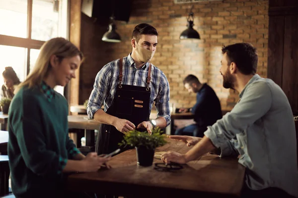 Happy waiter taking order and communicating with guests while wearing ...