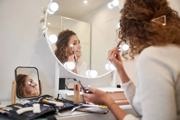 From below of reflection in mirror of young female doing facial makeup ...